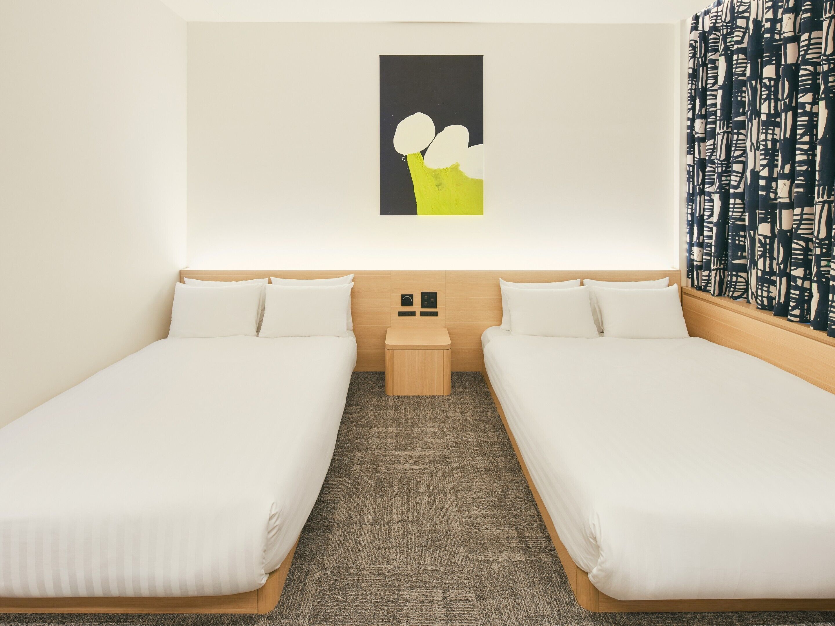 Quarto de hotel com duas camas de solteiro, uma pequena mesa de cabeceira, uma luminária fixada na parede e cortina estampada.
