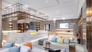 Um saguão de hotel moderno com uma recepção de mármore, área de estar com sofás, escadaria e uma tela de televisão.