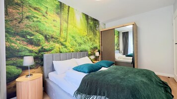 Quarto moderno com um grande mural de floresta, cama estofada cinza, almofadas verde-água e manta verde, além de uma mesinha de cabeceira de madeira com abajur.