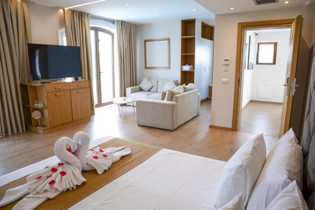 Quarto de hotel com cama, sofá, televisão, armário de madeira e uma janela grande.