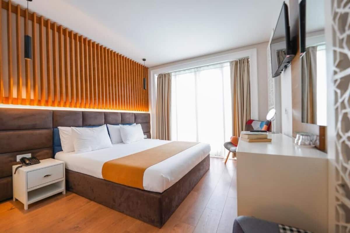 Quarto de hotel moderno com uma cama grande, uma mesa de cabeceira com telefone, uma escrivaninha com cadeira e uma TV na parede.