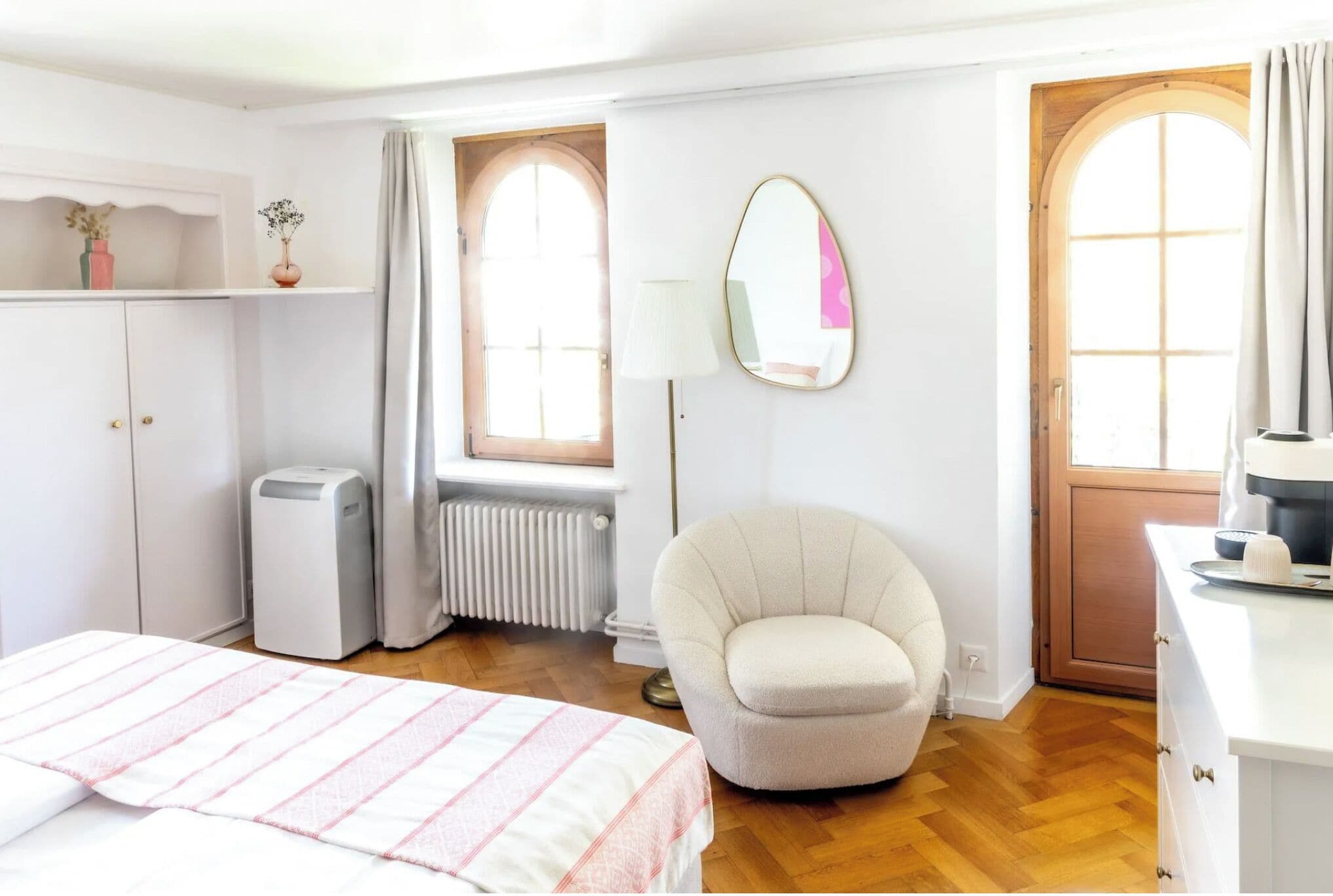 Um quarto com uma cama, uma cadeira, um radiador, um abajur, um espelho e duas janelas.
