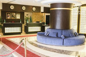 Um hall de hotel com uma mesa de recepção, um amplo sofá azul e um pilar decorativo.
