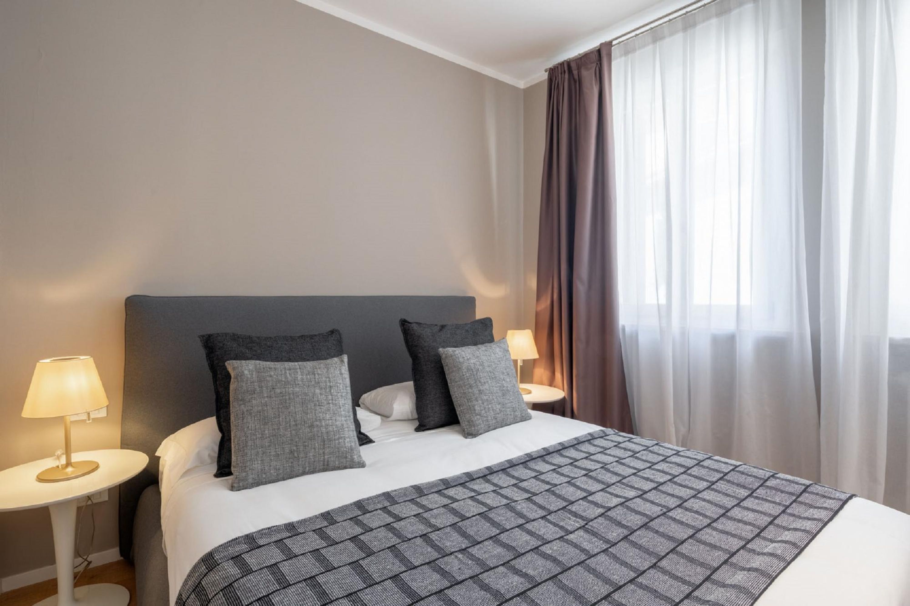 Uma cama bem arrumada com um cobertor xadrez, dois travesseiros e uma luminária de cabeceira em uma mesa de cabeceira branca.