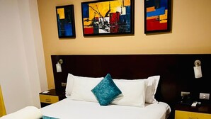 Quarto de hotel com uma cama grande, mesa de cabeceira e três quadros na parede.