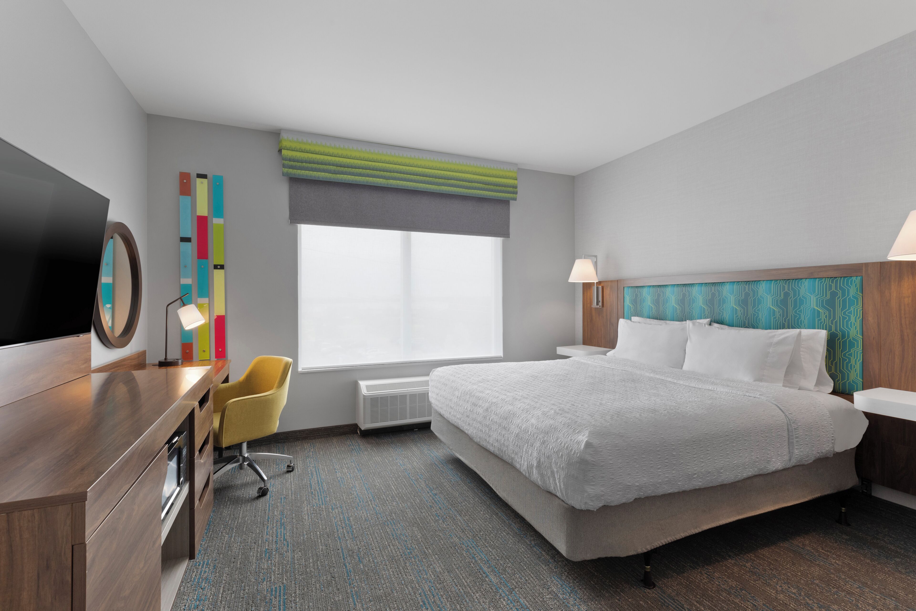 Quarto de hotel com uma cama grande, uma escrivaninha com cadeira, televisão, janela com persianas e um objeto decorativo colorido na parede.