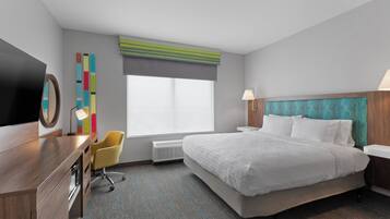 Quarto de hotel com uma cama grande, uma escrivaninha com cadeira, televisão, janela com persianas e um objeto decorativo colorido na parede.
