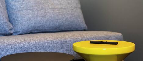 Um sofá com um travesseiro, uma mesinha redonda com superfície preta e um vaso amarelo com um objeto preto em cima.