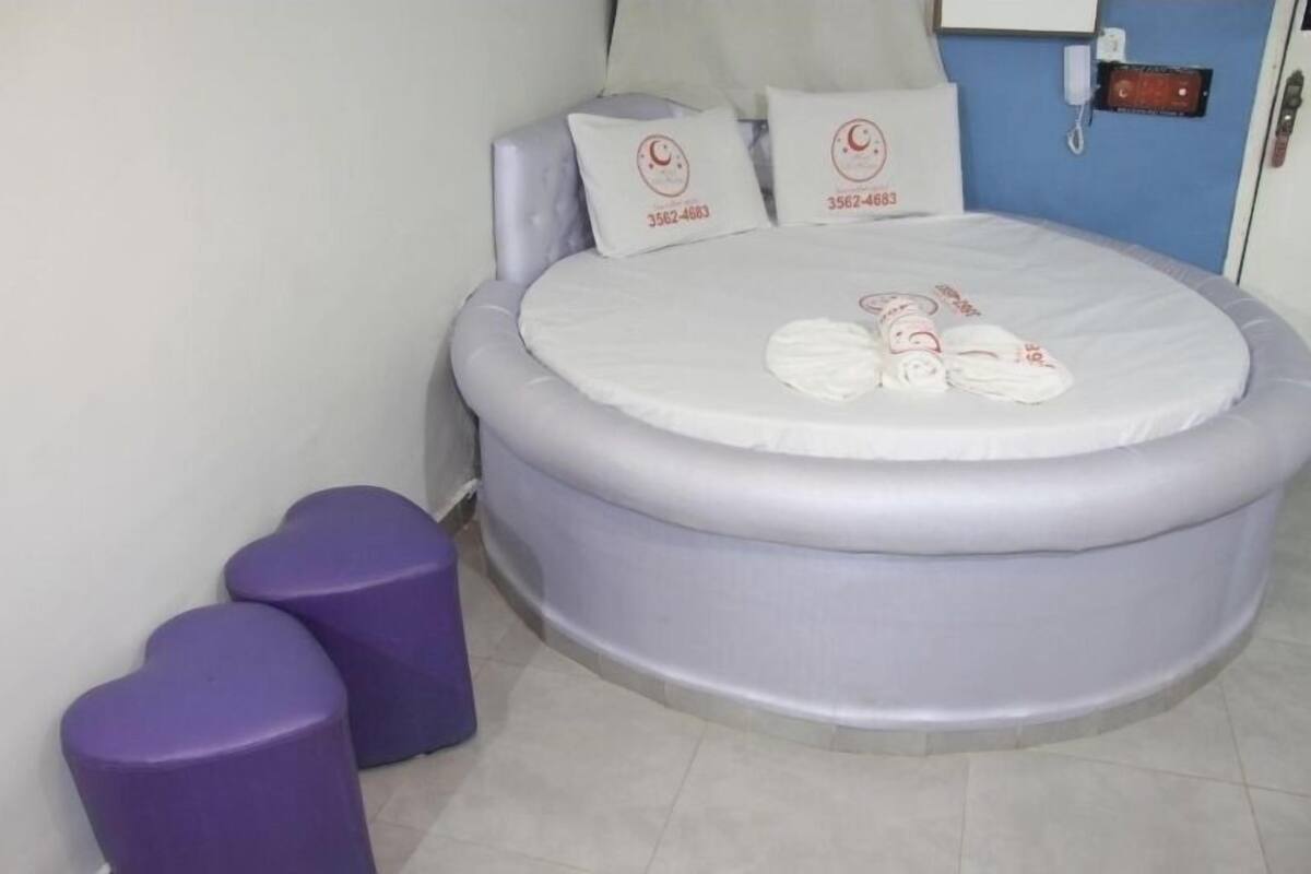 Uma cama redonda com colchão e almofadas brancas, dois banquinhos roxos e um telefone na parede.