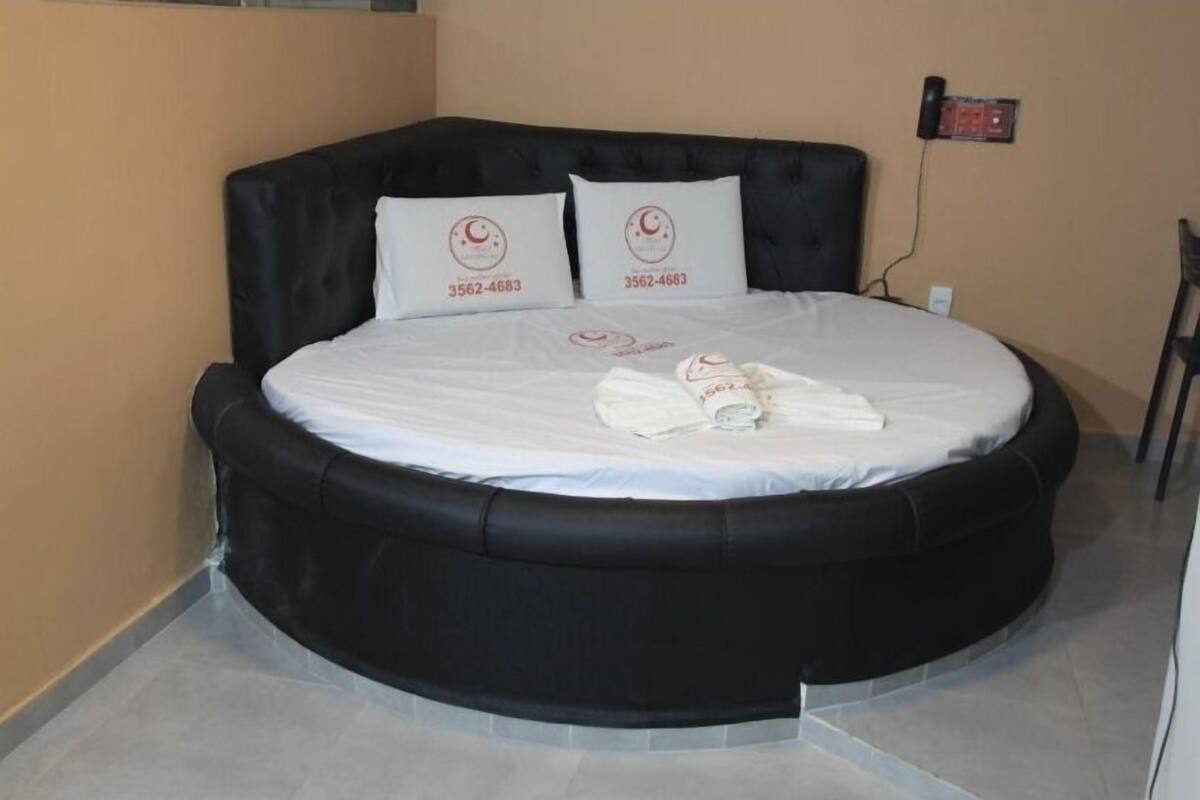 Cama redonda com cabeceira preta e duas almofadas brancas com logotipo e número. Há toalhas sobre a cama.