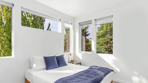 Quarto com cama grande, paredes brancas e três janelas com vista para árvores.