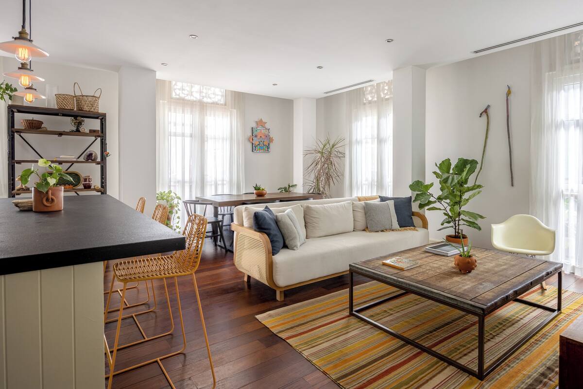 Uma sala de estar moderna com sofá, mesa de centro e cadeiras. Há uma área de jantar com mesa e cadeiras. O ambiente possui janelas amplas com cortinas leves.