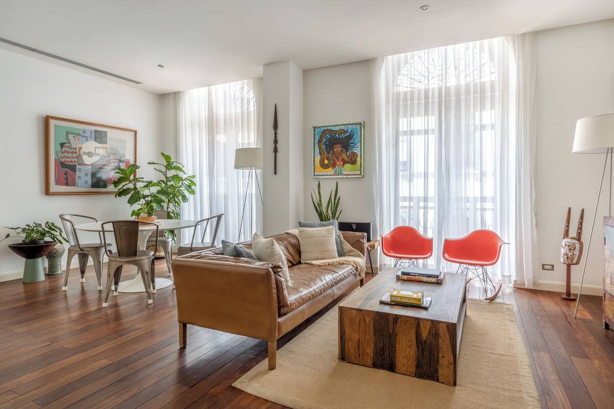 Uma sala de estar moderna com um sofá de couro, uma mesa de centro de madeira e cadeiras vermelhas. Há plantas em vasos, um quadro e um abajur sobre uma mesinha lateral.