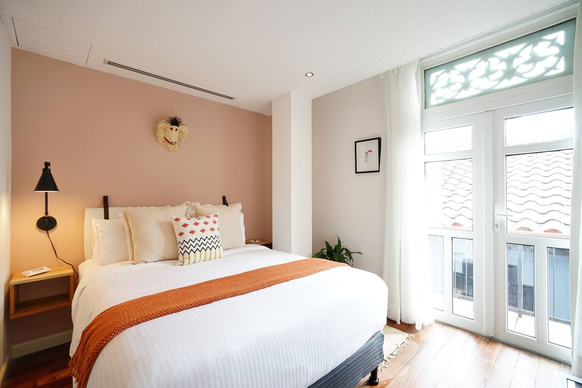 Quarto com uma cama grande, uma mesinha de cabeceira, uma luminária na parede, um objeto decorativo na parede e vista para o exterior através de uma porta de vidro.