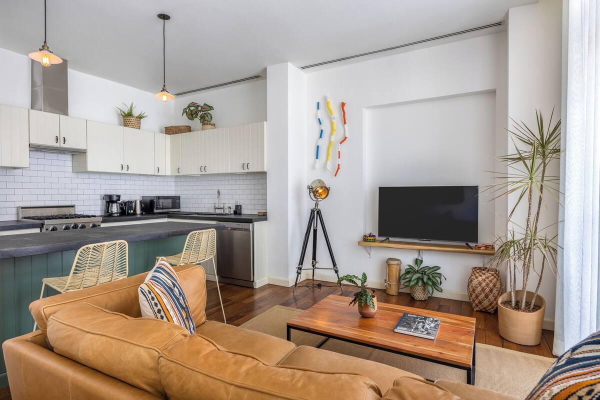 Uma sala moderna com um sofá de couro, uma mesa de centro e uma TV sobre um suporte. Há uma área de cozinha com armários brancos e bancada preta.