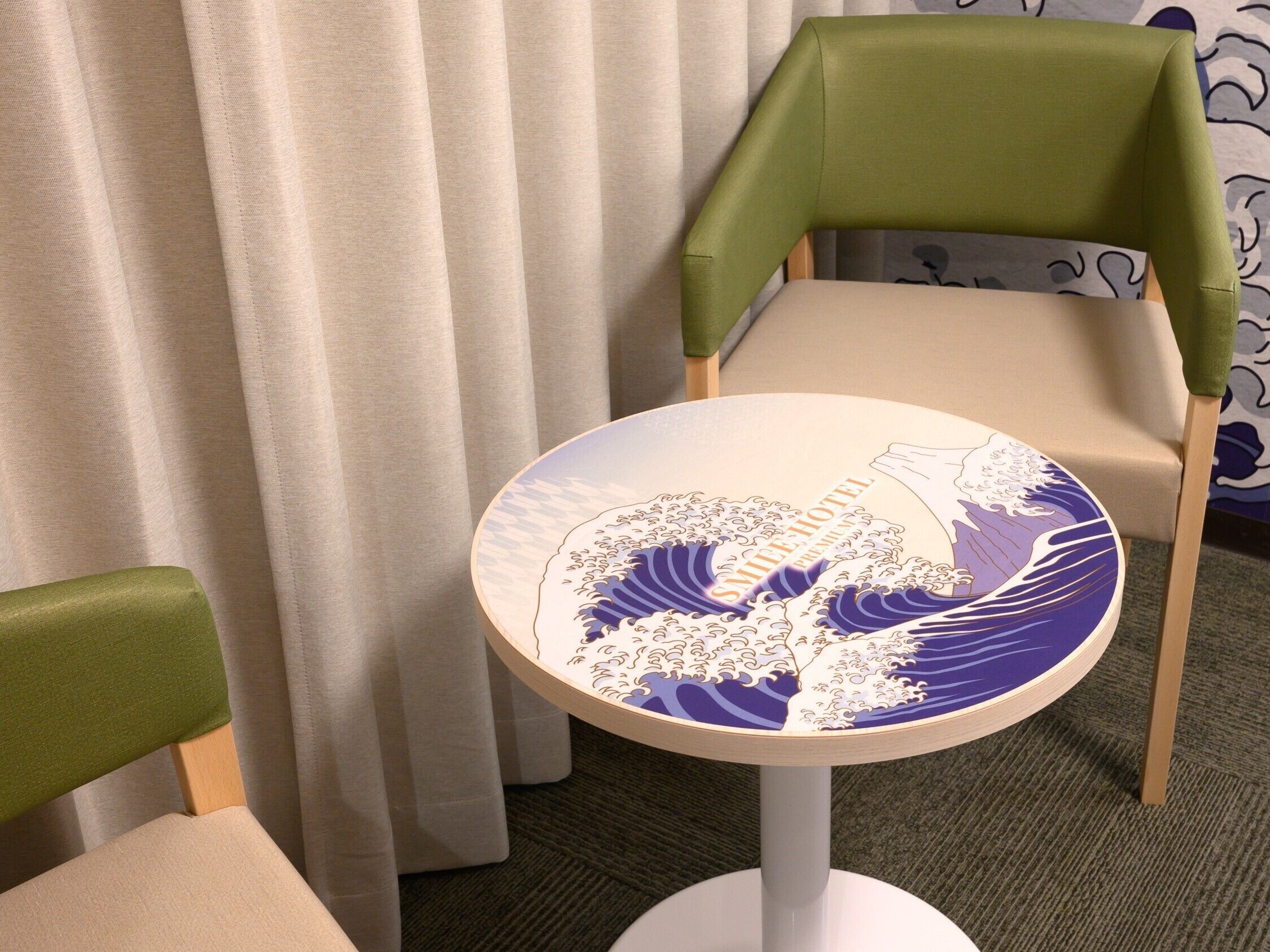 Uma mesa redonda com design de onda japonesa, uma cadeira estofada verde e uma cadeira estofada bege em um quarto com cortina e papel de parede estampado.