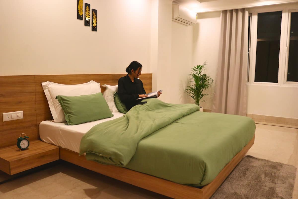 Cama bem arrumada com manta verde, mesa de cabeceira com relógio e planta em vaso perto da janela.