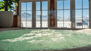 Área de spa com piso aquecido verde, uma planta em vaso e janelas amplas com vista para as montanhas.