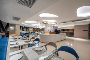 Uma área de refeições moderna, com mesas preparadas para refeições, assentos azuis e um balcão central.