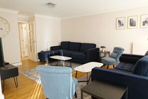 Uma sala de estar com um sofá azul escuro, uma poltrona azul clara, uma mesa de centro branca e uma mesinha lateral.