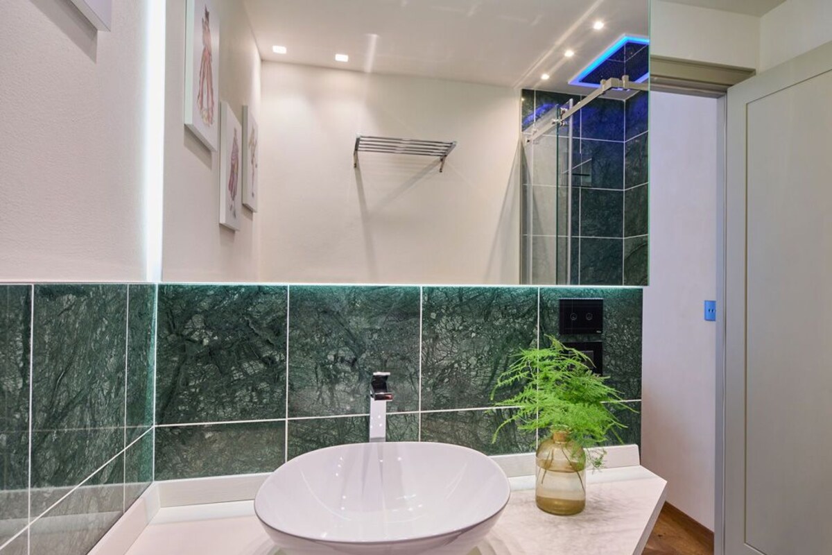 Casa de banho com lavatório embutido branco, azulejos de mármore verde, cabine de duche em vidro e uma pequena planta no balcão.