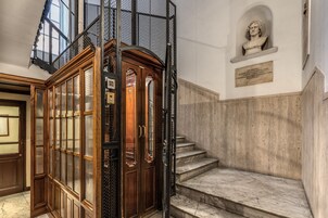 Uma escadaria de mármore com elevador de madeira e um busto na parede.