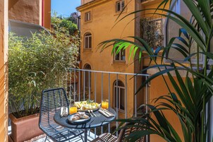Uma varanda com uma mesa posta para o café da manhã, incluindo croissants e suco de laranja, cercada por plantas e um edifício amarelo.