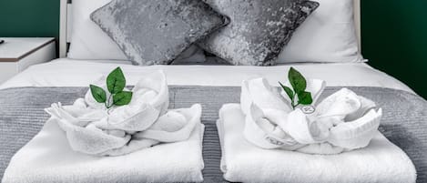 Cama bem arrumada com lençóis brancos e duas toalhas com folhas verdes, um cobertor cinza e duas almofadas cinzas em um fundo de parede verde.