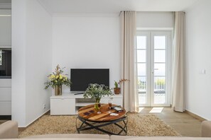 Uma sala de estar moderna com uma televisão de tela plana, uma mesa de centro de madeira e um sofá. Há uma janela grande com cortinas, um vaso de planta e uma pequena mesa com bebidas e petiscos.