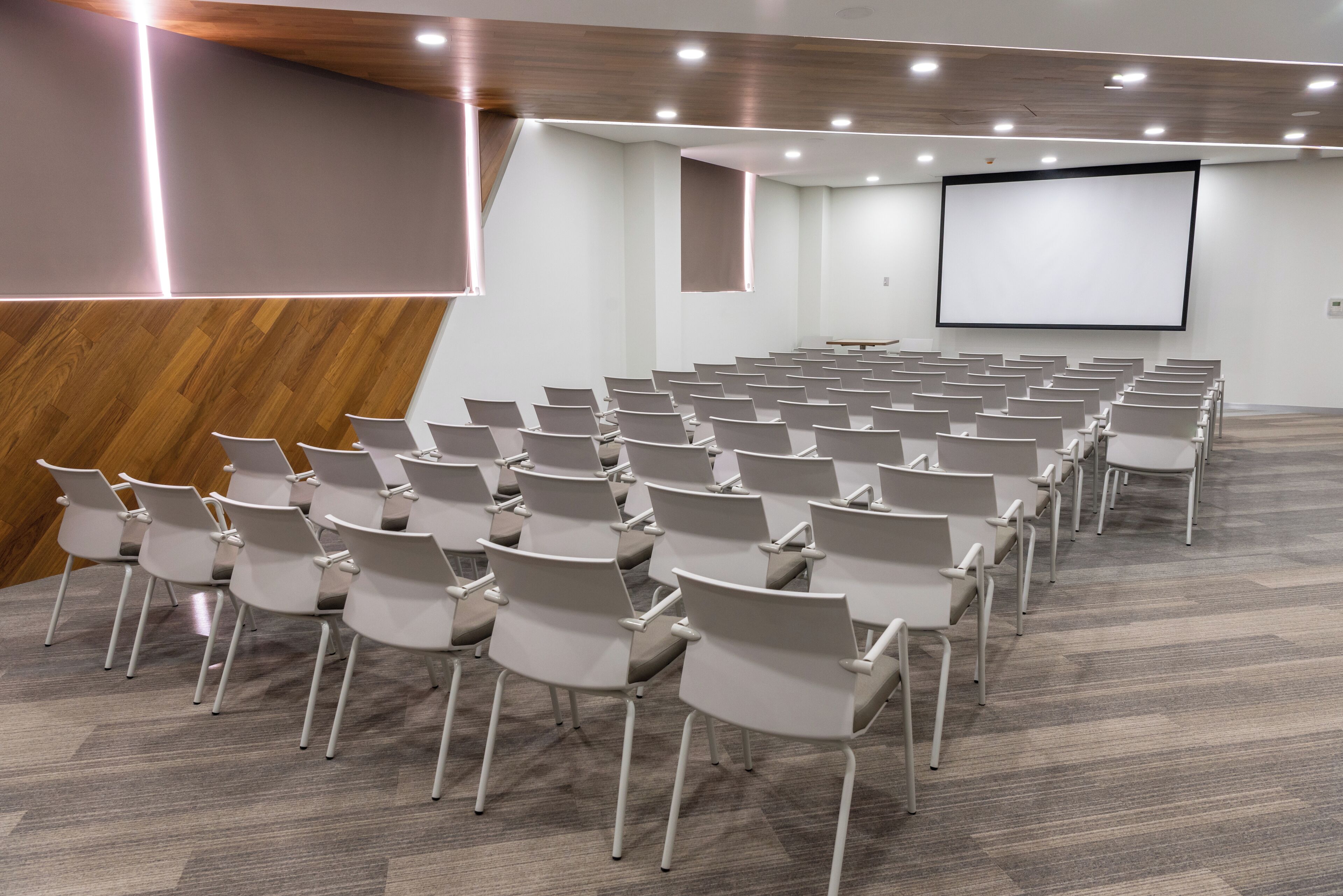 Sala de conferências com fileiras de cadeiras brancas, uma tela grande e uma parede de madeira decorativa.
