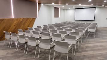 Sala de conferências com fileiras de cadeiras brancas, uma tela grande e uma parede de madeira decorativa.