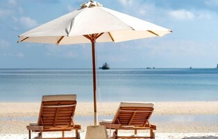 Duas cadeiras de madeira para relaxar sob um guarda-sol branco numa praia, com um mar calmo e um navio ao longe.