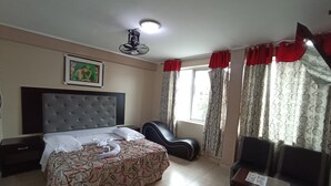Quarto de hotel com cama, poltrona, ventilador, janela com cortinas e um quadro na parede.