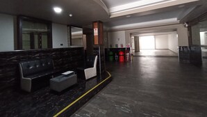 Um lobby espaçoso com um balcão de recepção em mármore preto, área de estar com um banco, e vista desobstruída para o estacionamento ao fundo.