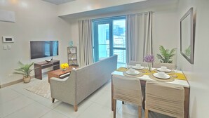 Sala de estar moderna com sofá, mesa de jantar e cadeiras. Há uma televisão de tela plana na parede, uma mesinha de apoio com abajur e um vaso de planta.