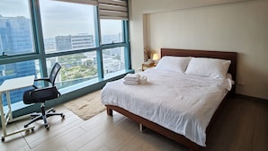 Quarto de hotel moderno com uma grande janela, cama de madeira, escrivaninha com cadeira e vista para a paisagem urbana.