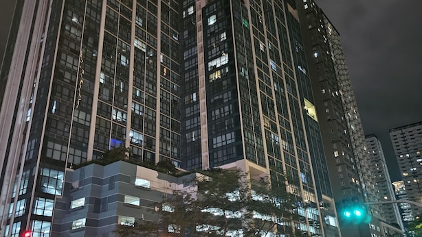 Um edifício alto com vários andares e grandes janelas, uma rua com veículos e pedestres, e semáforos.