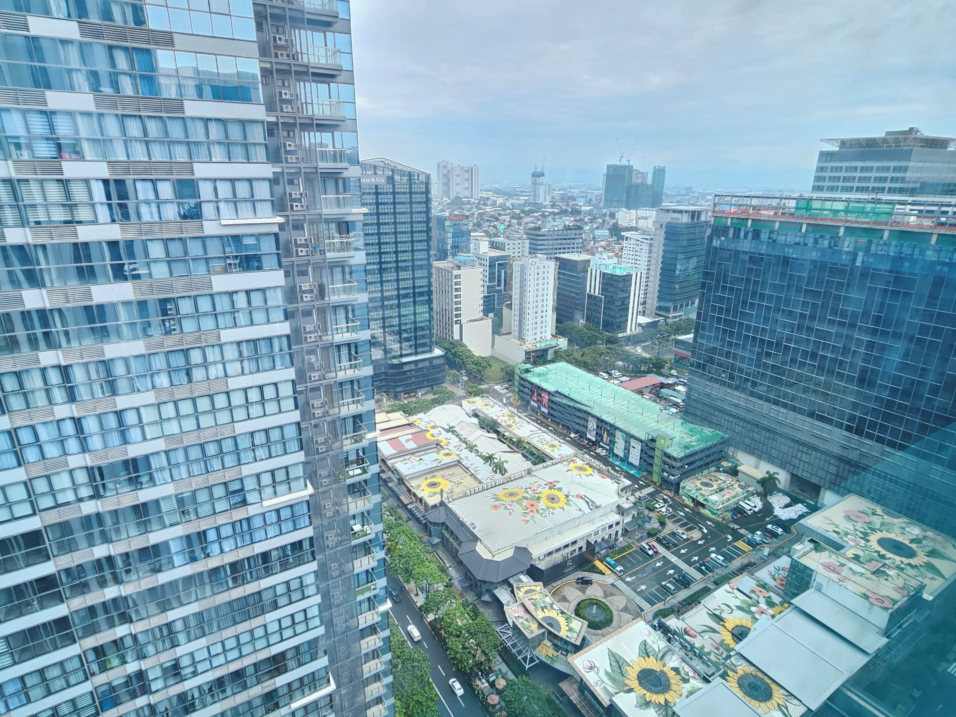 Um edifício alto com fachada de vidro moderna, cercado por outros edifícios altos e uma praça central com telhado verde e murais de girassóis.