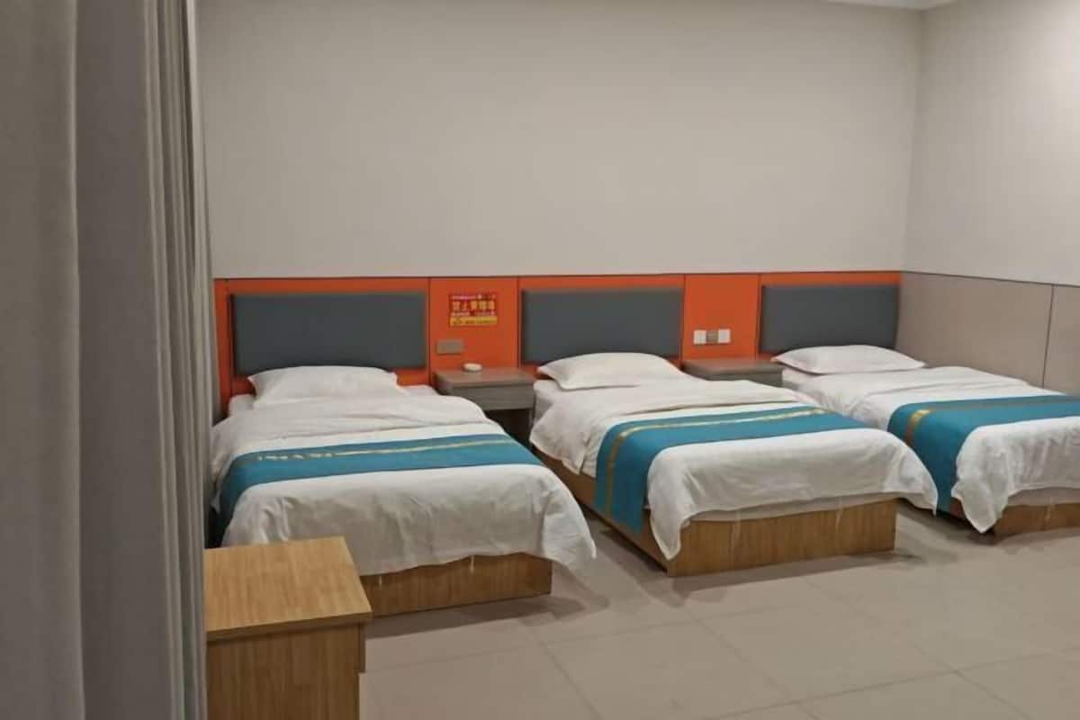 Quarto de hotel com três camas individuais, cada uma com cabeceira de madeira e roupa de cama branca. Há uma pequena mesa de cabeceira de madeira entre as camas.