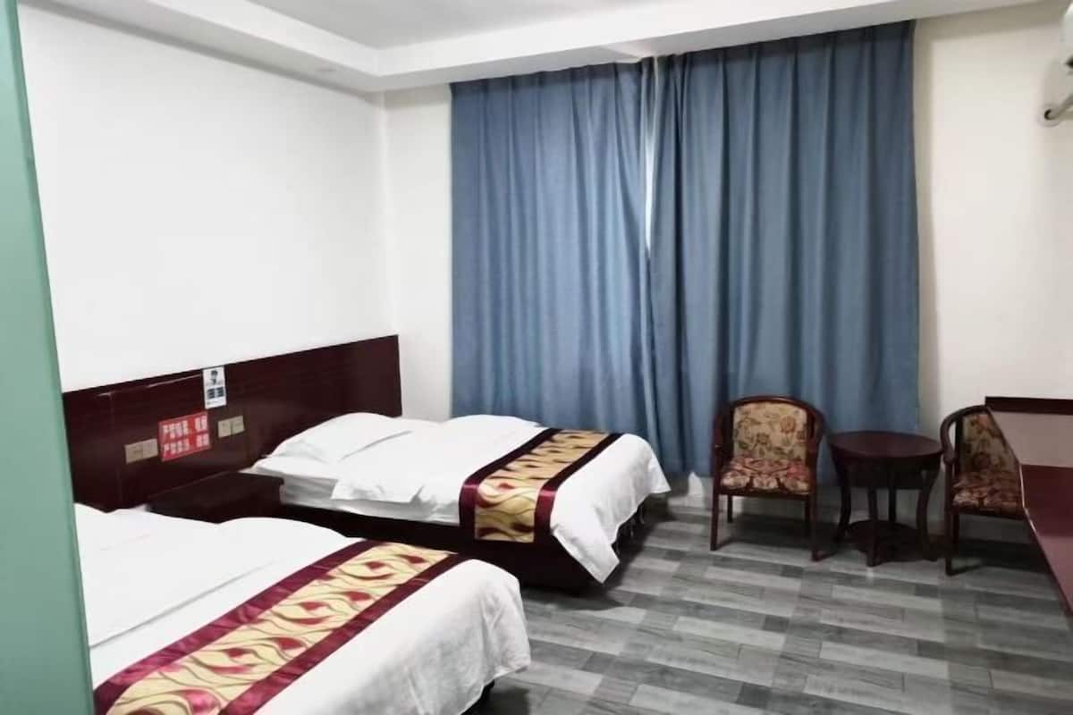 Quarto de hotel com duas camas, uma mesa pequena, duas cadeiras e uma janela grande com cortinas azuis.