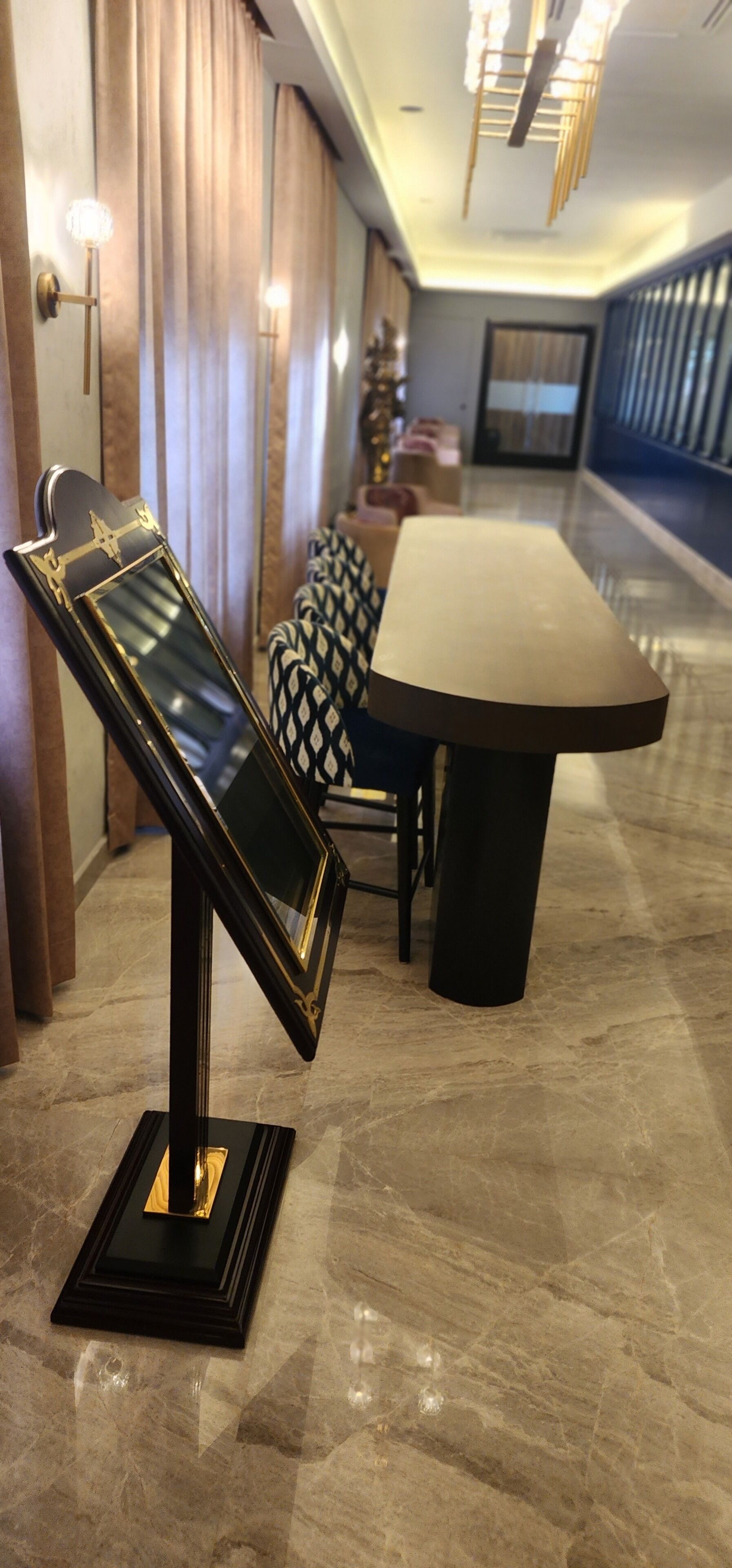 Um hall de hotel moderno com piso de mármore, um espelho com moldura preta e dourada, uma mesa de jantar e uma cadeira estampada.