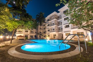 Uma piscina rodeada por um edifício com varandas, palmeiras e céu noturno.