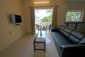 Uma sala de estar com sofá, mesa de centro, televisão e varanda com vista para edifícios e árvores.