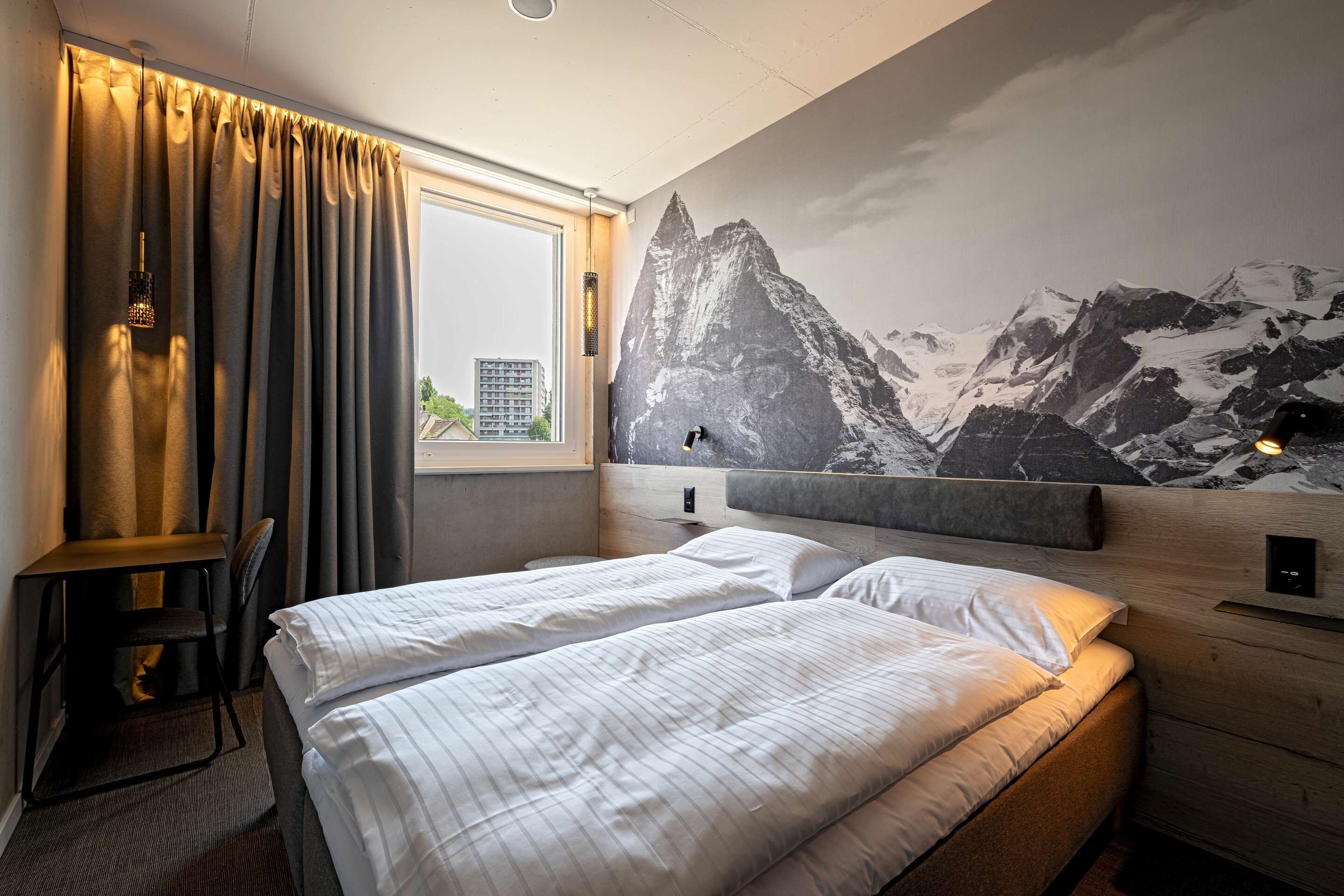 Quarto de hotel com uma cama grande, uma cadeira, uma mesa e uma janela com cortinas.