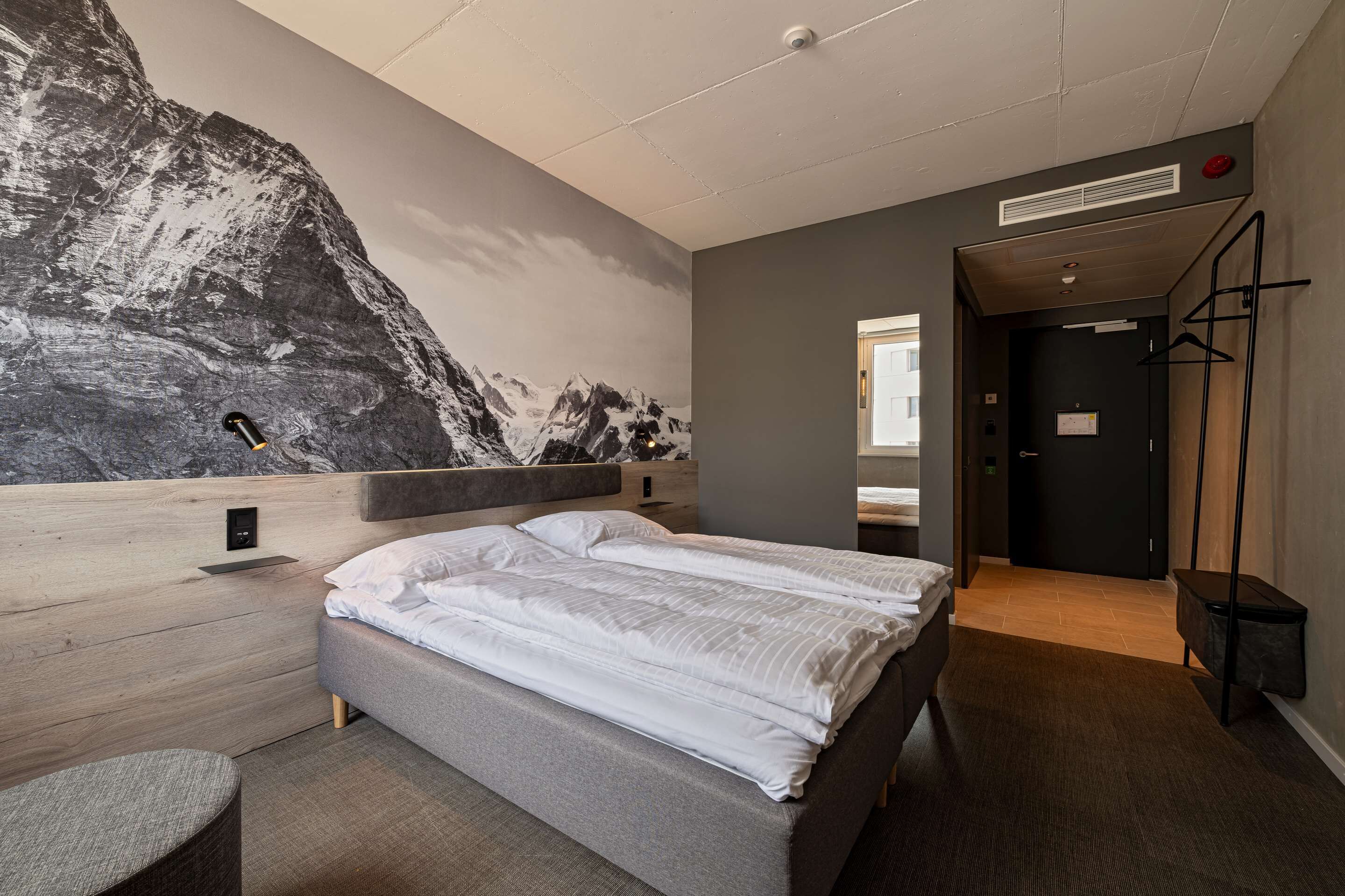 Quarto de hotel moderno com um grande mural de montanhas na parede, uma cama com roupa de cama branca, uma mesa de cabeceira e um pequeno espaço de estar circular.