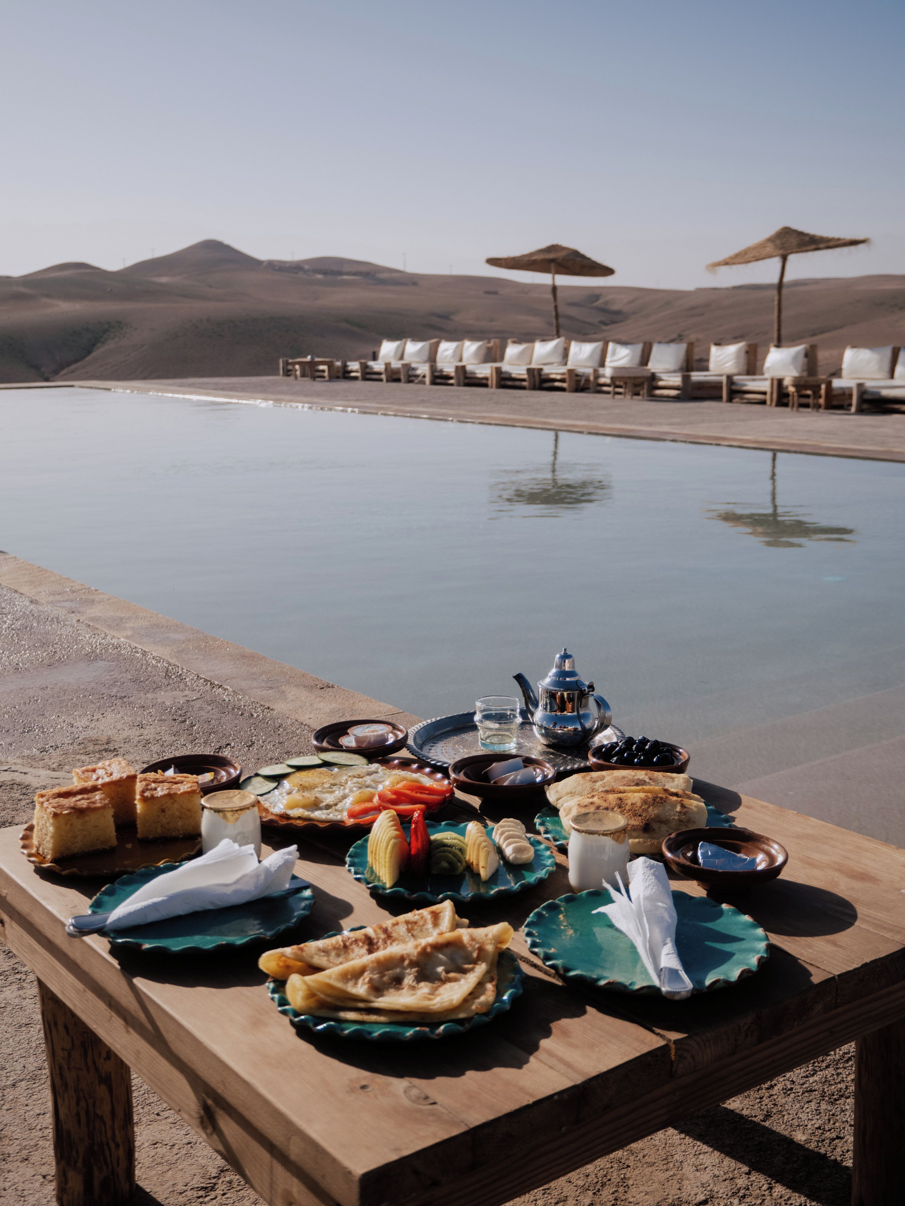 Mesa de jantar com diversos pratos e um bule junto a uma piscina, com cadeiras de sol e guarda-sóis ao fundo.