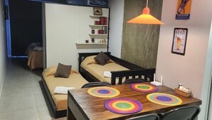 Um espaço de estar compacto com uma cama, um sofá pequeno, uma mesa de jantar com suportes de copo coloridos, um abajur e uma estante de livros.