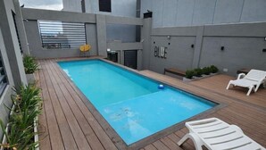 Uma área moderna de piscina ao ar livre com um deque de madeira, cadeiras de descanso e um edifício com um letreiro 'BLUE'.