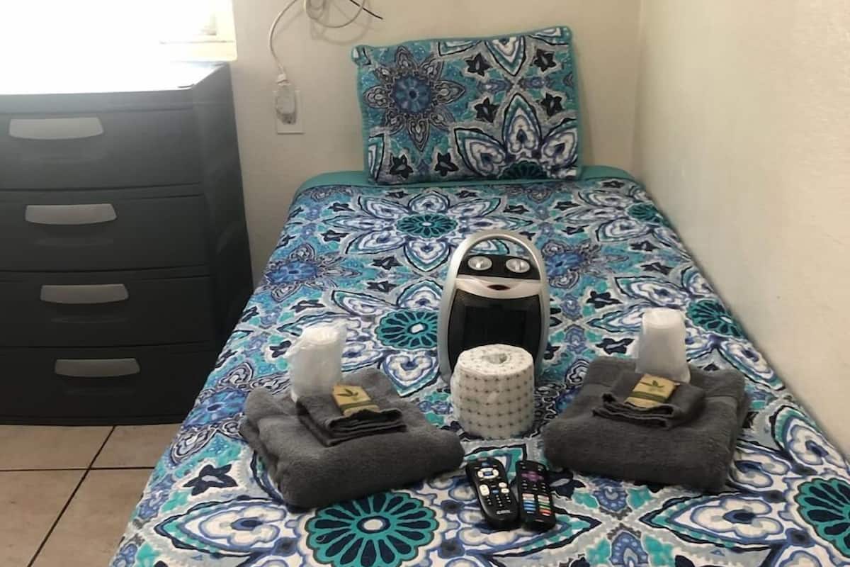 Um quarto pequeno e bem arrumado com colcha estampada, um travesseiro, mesa de cabeceira com abajur e ar condicionado fixado na parede.