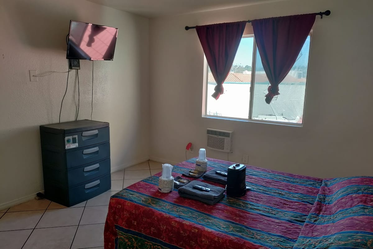Quarto com cama, penteadeira, TV fixada na parede, janela com cortinas e ar condicionado.